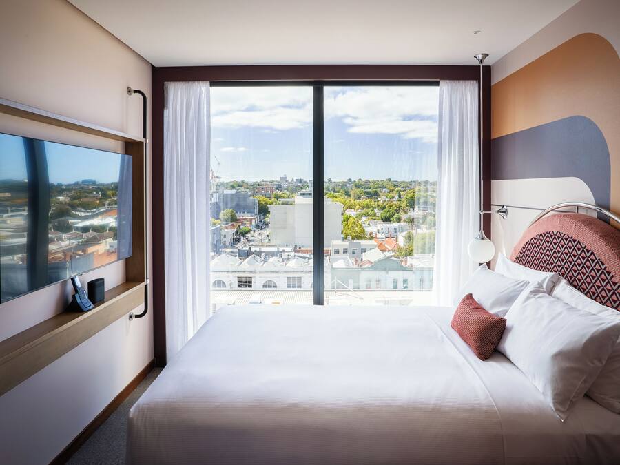Quarto de hotel com uma cama grande, televisão de tela plana na parede, janela com vista para a paisagem urbana e cabeceira moderna.