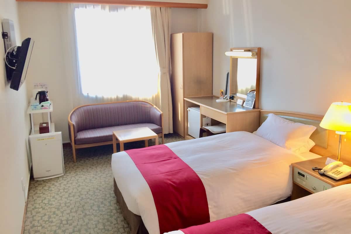 Quarto de hotel com duas camas, uma escrivaninha, uma cadeira, uma mesinha, um espelho, um abajur, um telefone e uma janela com cortinas.