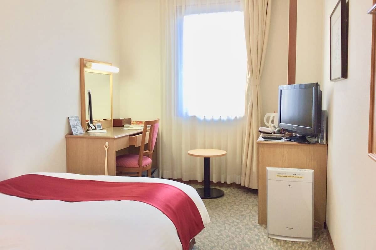 Quarto de hotel com cama, escrivaninha com cadeira, televisão e uma mesinha.