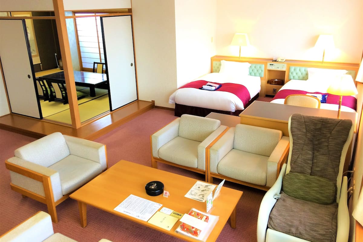 Quarto de hotel com uma cama, duas poltronas, uma mesa de centro com papéis, uma escrivaninha e um espelho.