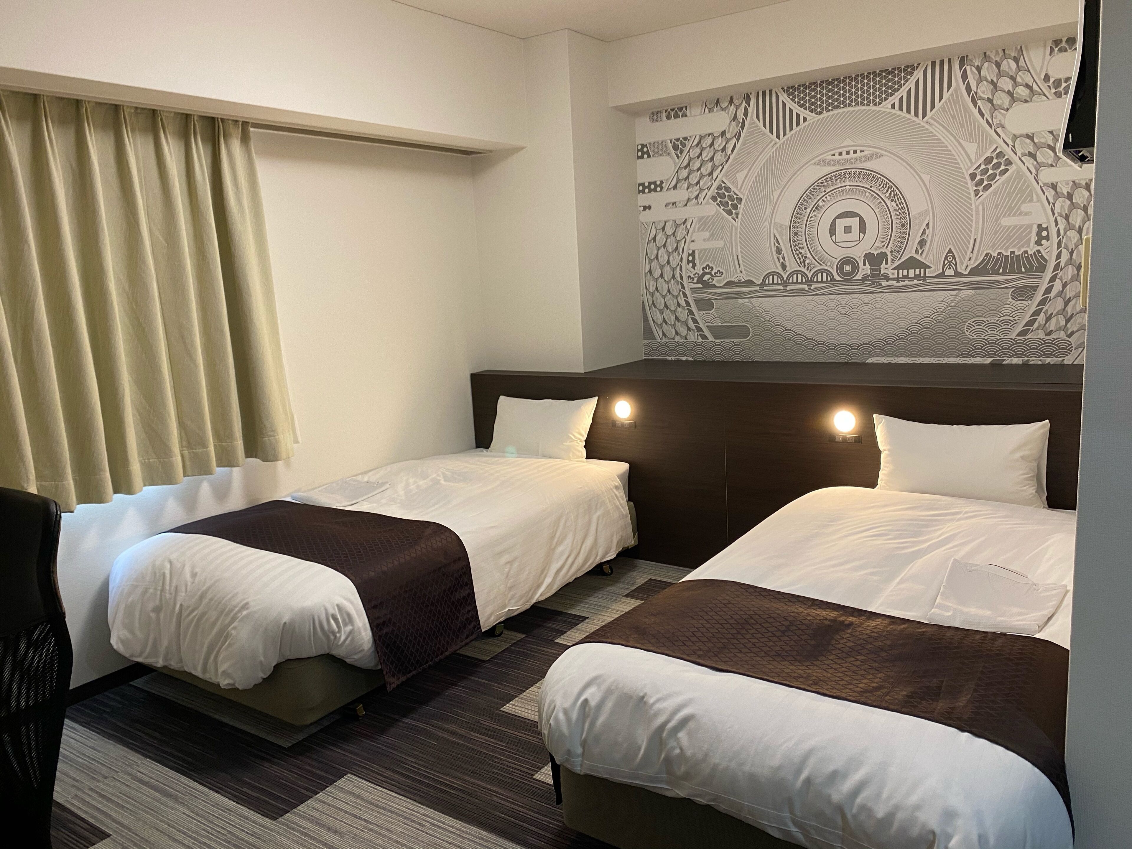 Quarto de hotel com duas camas, cabeceira com iluminação integrada, um grande tapeçaria na parede e uma janela com cortinas.