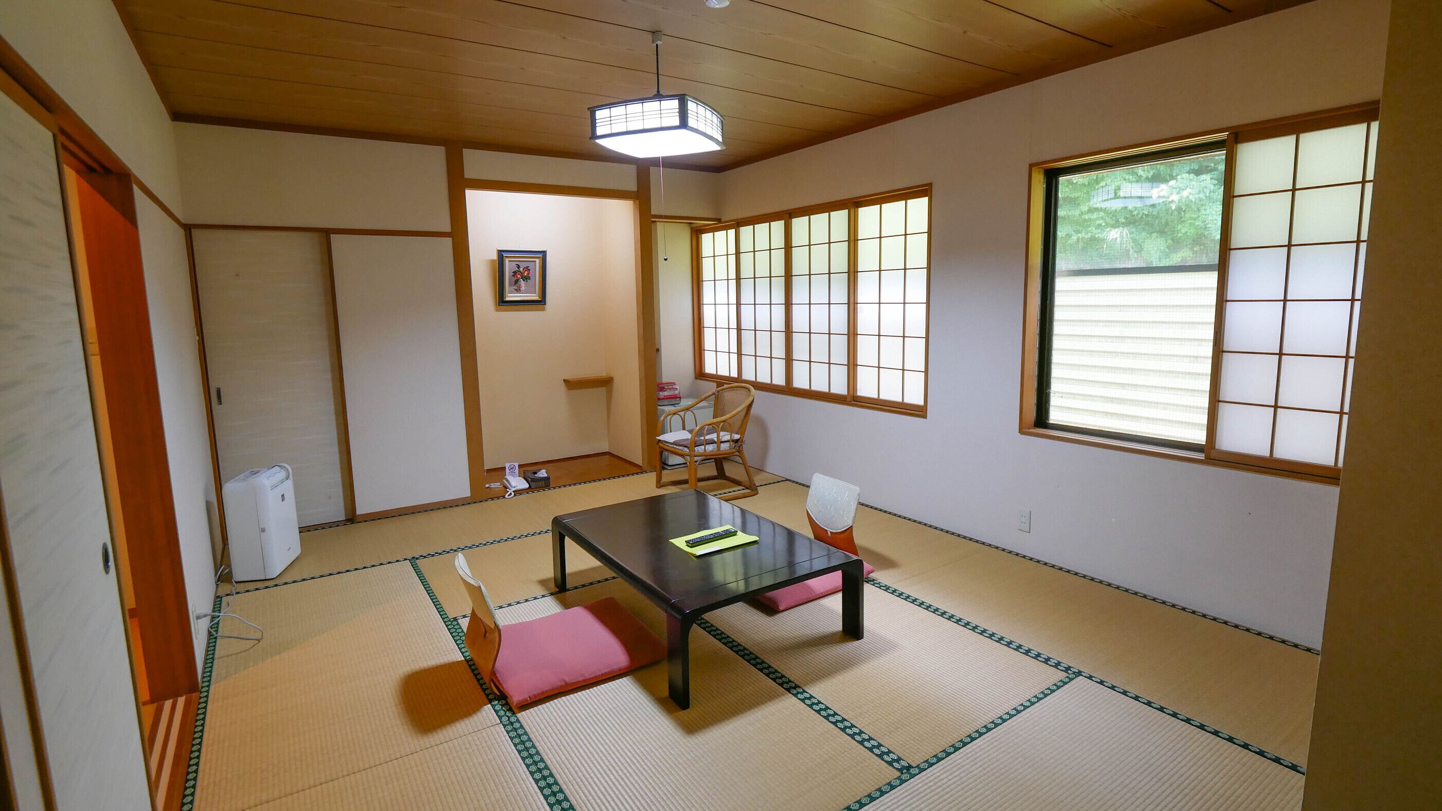 Um quarto japonês tradicional com tatames, uma mesa baixa com almofadas, uma cadeira, uma janela com biombos shoji e um ventilador de teto.