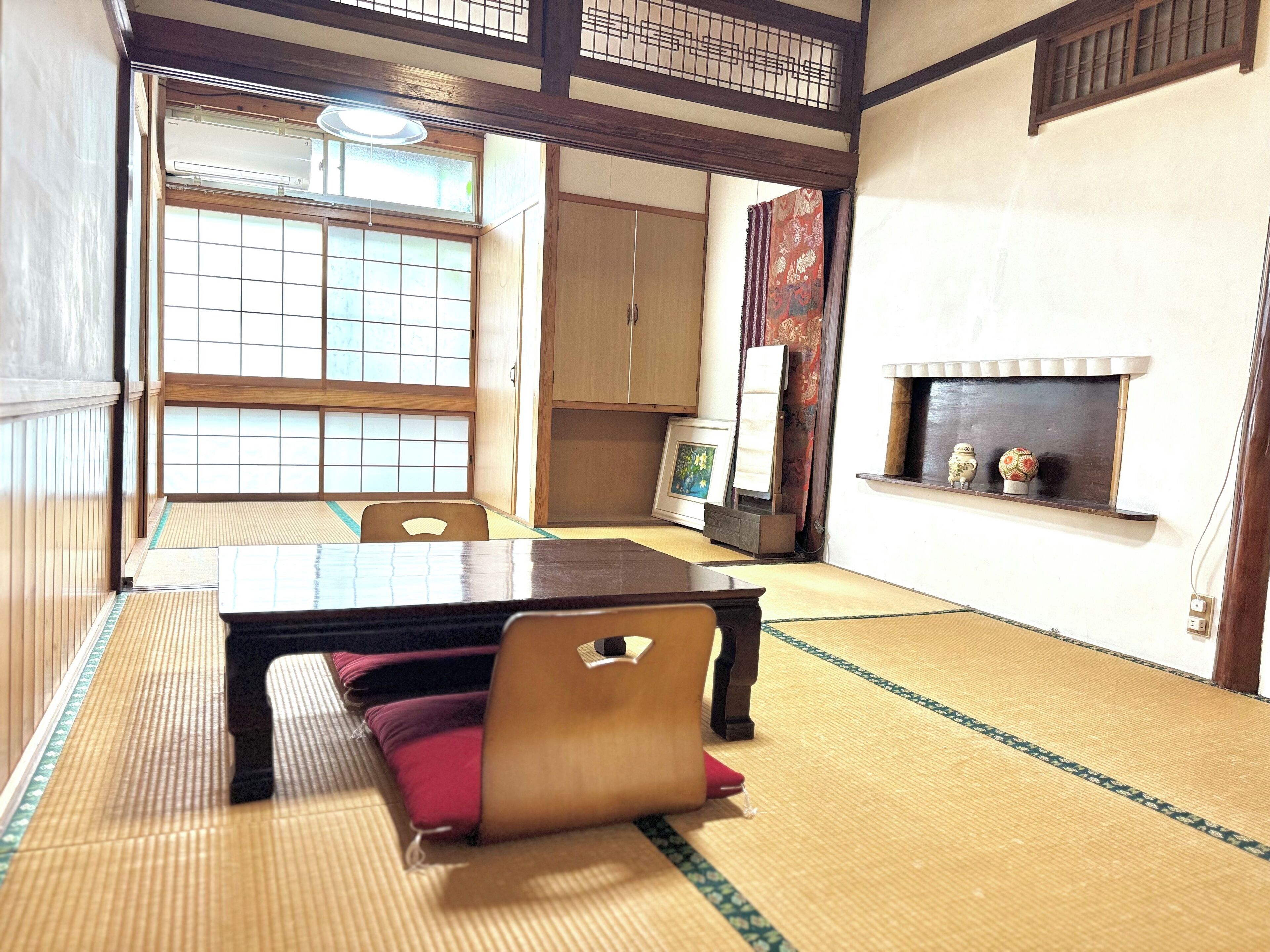 Um espaço com tatames, uma mesa baixa, um banco acolchoado, uma porta deslizante, uma estante japonesa decorada com objetos e uma obra de arte emoldurada.