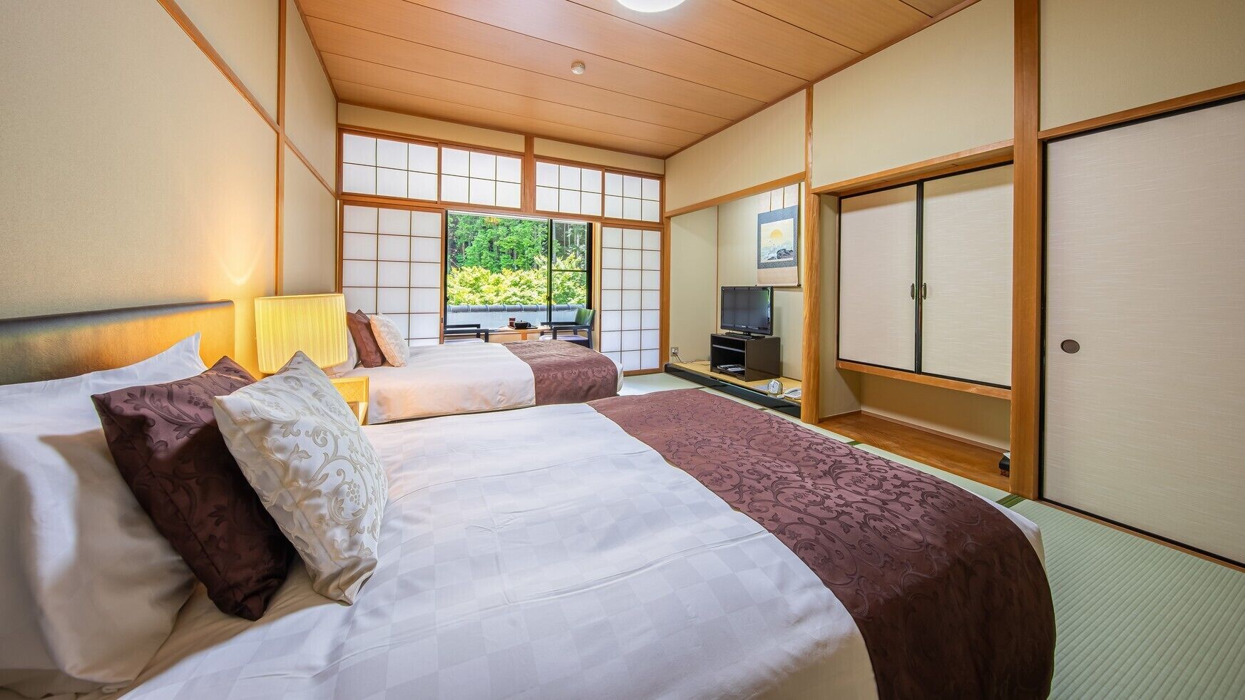 Quarto de hotel em estilo japonês tradicional, com piso de tatame, duas camas com roupa de cama branca e castanha, uma televisão e uma porta deslizante que dá para uma varanda com vista para o verde.