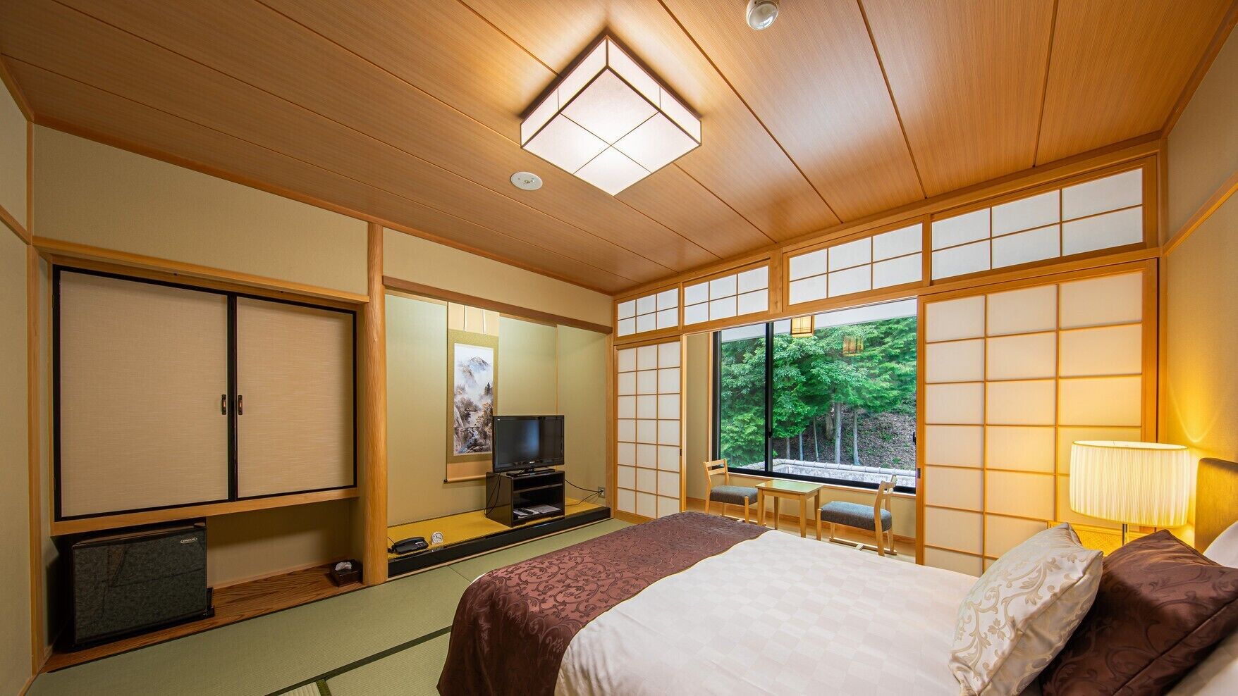 Quarto tradicional em estilo japonês com tatames, uma cama, televisão, escrivaninha e janela com vista para o verde.