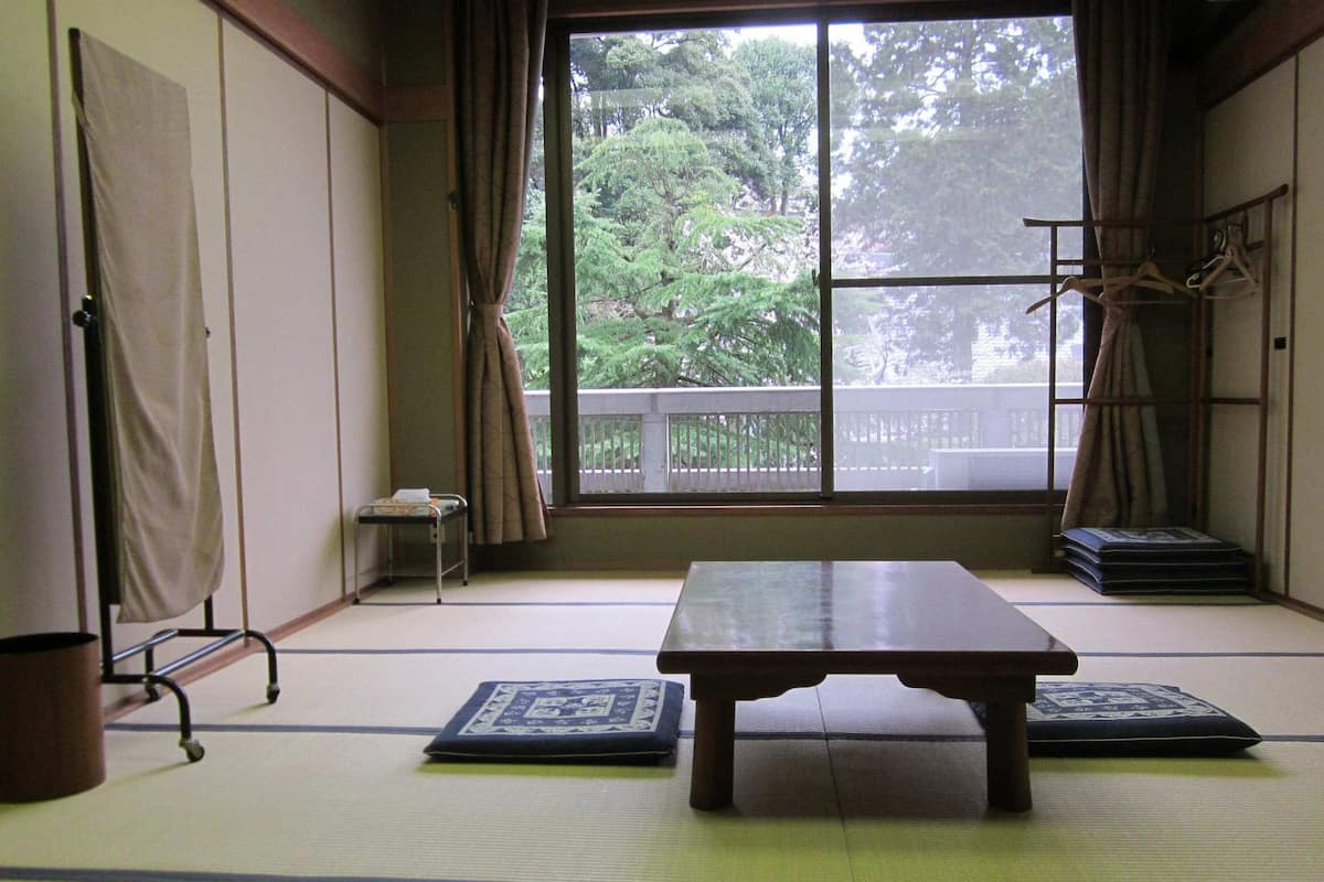 Um quarto japonês tradicional com piso de tatame, uma mesa de madeira baixa e vista para uma varanda com vegetação.