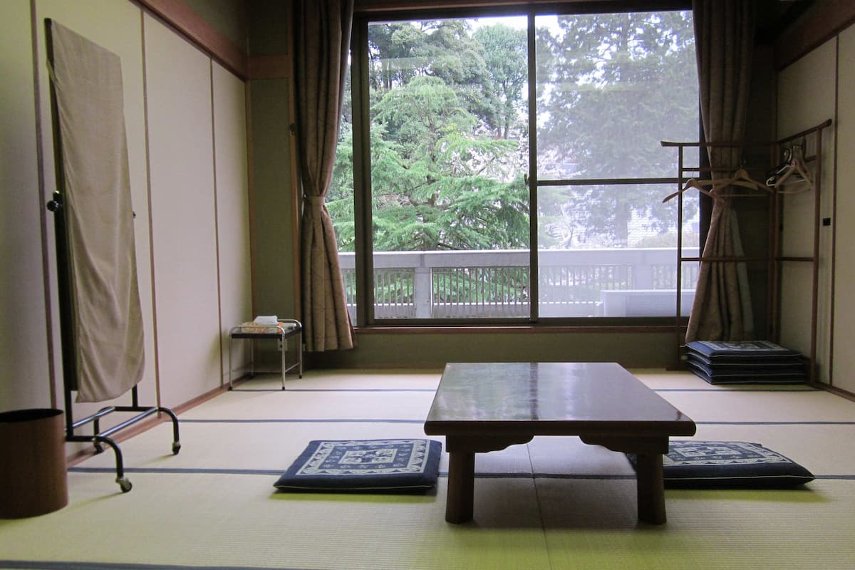 Um quarto japonês tradicional com piso de tatame, uma mesa baixa e vista para uma varanda com vegetação.