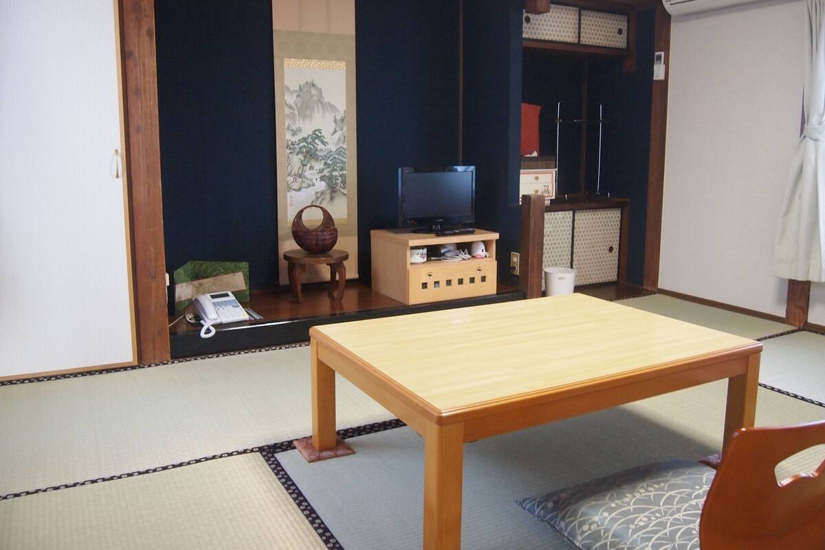 Um quarto japonês tradicional com tatames, uma mesa de madeira, uma televisão sobre um suporte e uma porta de correr.