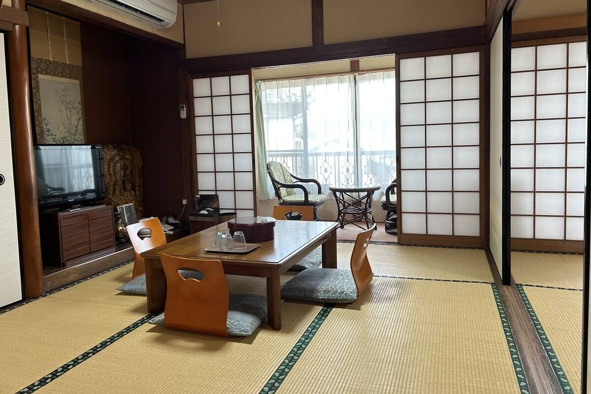 Um quarto japonês tradicional com piso de tatame, uma mesa de tatame, mobiliário de madeira, uma televisão e portas corrediças.