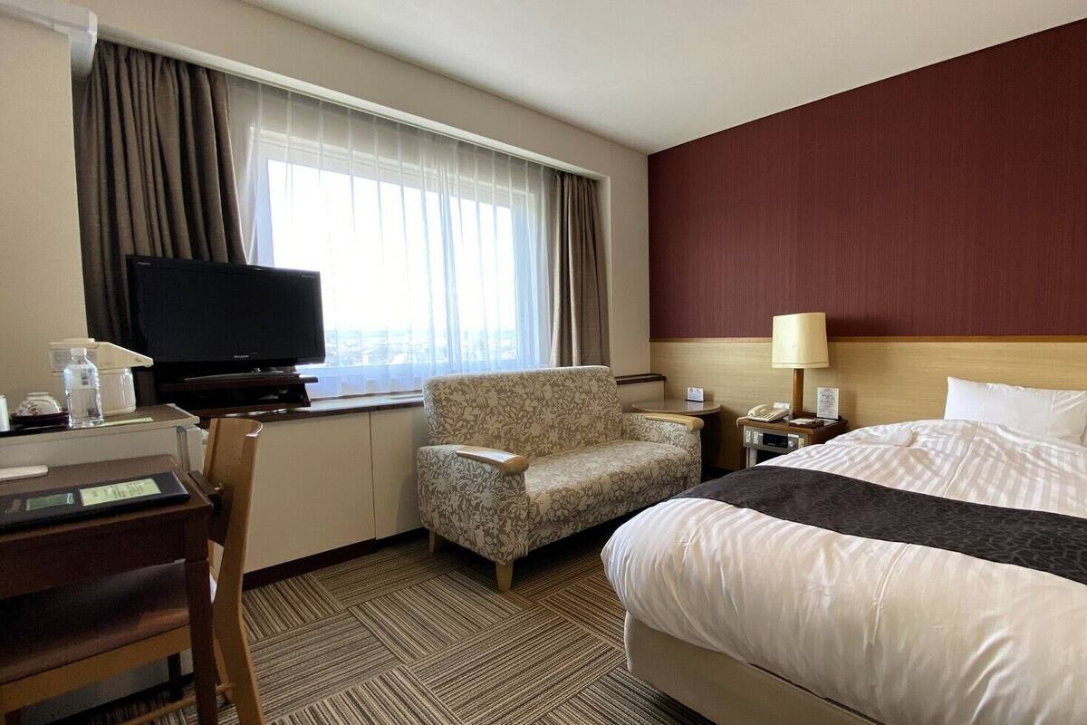 Quarto de hotel com cama, escrivaninha, cadeira, televisão e janela com cortinas.