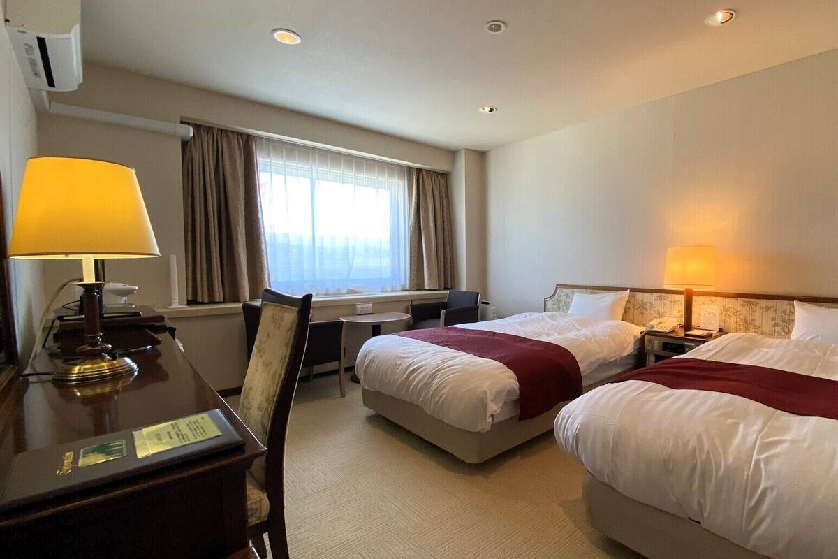 Quarto de hotel com duas camas, uma escrivaninha, uma cadeira, um abajur, uma janela com cortinas e um aparelho de ar condicionado.