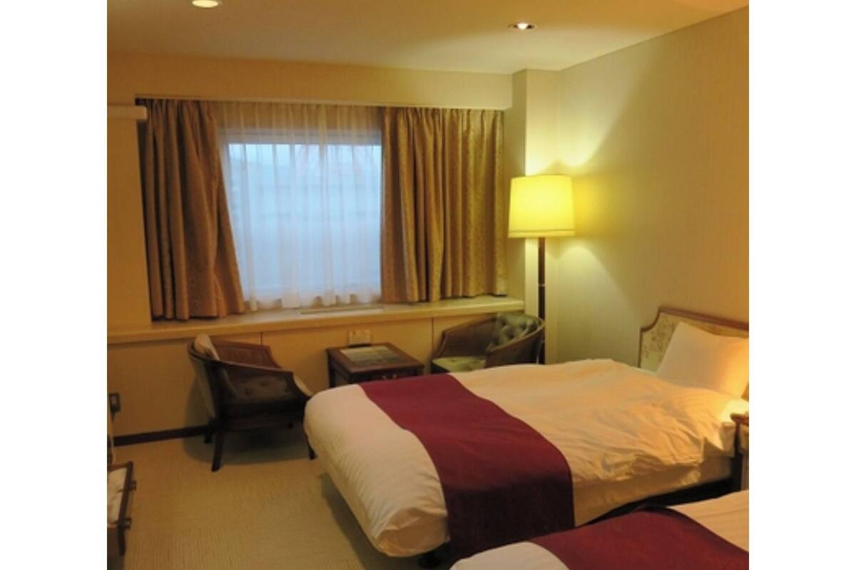 Quarto de hotel com duas camas, uma escrivaninha, uma cadeira, um abajur e uma janela com cortinas.