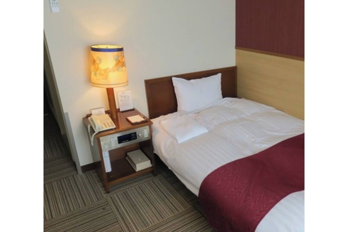 Quarto de hotel com cama bem arrumada, abajur, telefone e mesinha com um livro e um bloco de anotações.