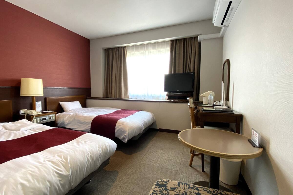 Quarto de hotel com duas camas, uma escrivaninha, televisão, espelho e aparelho de ar condicionado.
