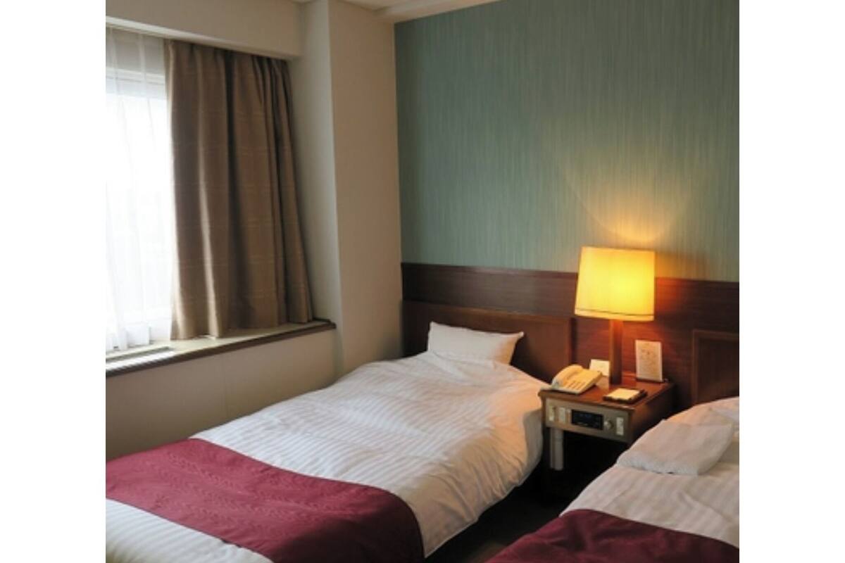 Quarto de hotel com duas camas, mesa de cabeceira com abajur e janela com cortinas.