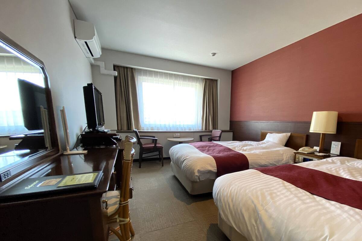 Quarto de hotel com duas camas, uma escrivaninha, televisão, uma cadeira e janela com cortinas.