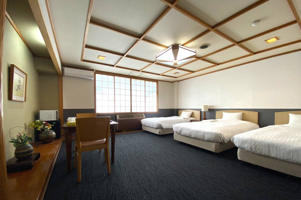 Quarto de hotel com escrivaninha, duas camas, uma cadeira e uma janela grande.