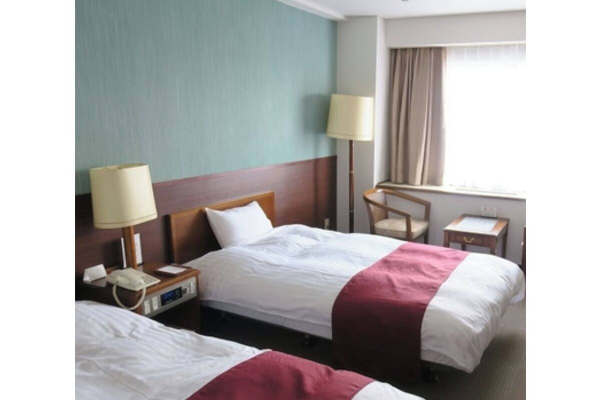 Quarto de hotel com duas camas, uma escrivaninha, uma cadeira, um abajur e uma janela com cortinas.