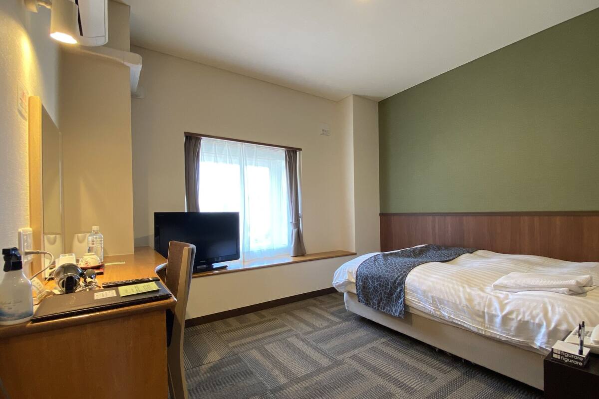 Quarto de hotel com cama, escrivaninha com computador, cadeira, televisão e janela com cortinas.