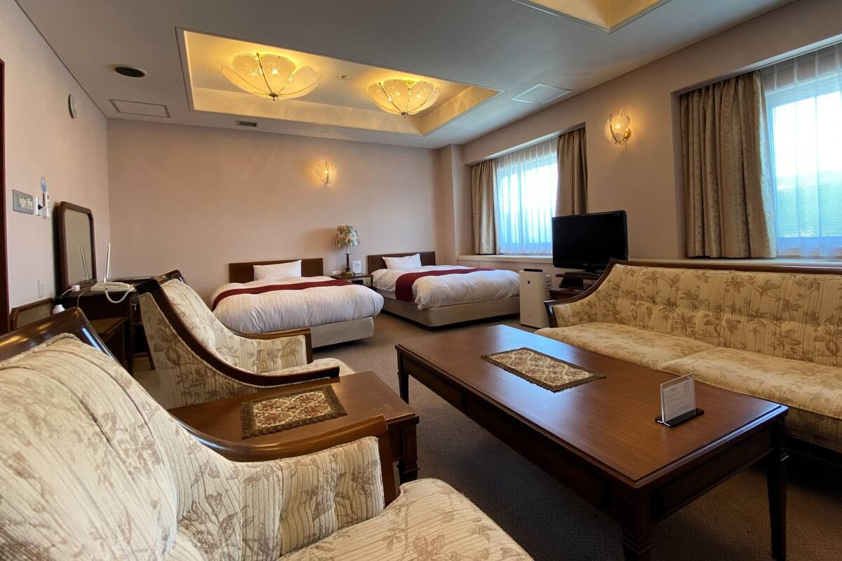 Quarto de hotel com cama, duas poltronas, um sofá, mesa de centro, televisão e uma janela grande com cortinas.