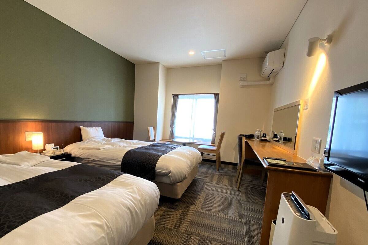 Quarto de hotel com duas camas, uma mesa de trabalho com uma TV, uma cadeira e uma janela.