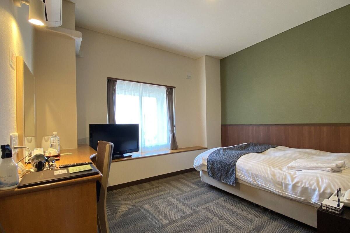 Quarto de hotel com cama, escrivaninha com computador, televisão e janela com cortinas.