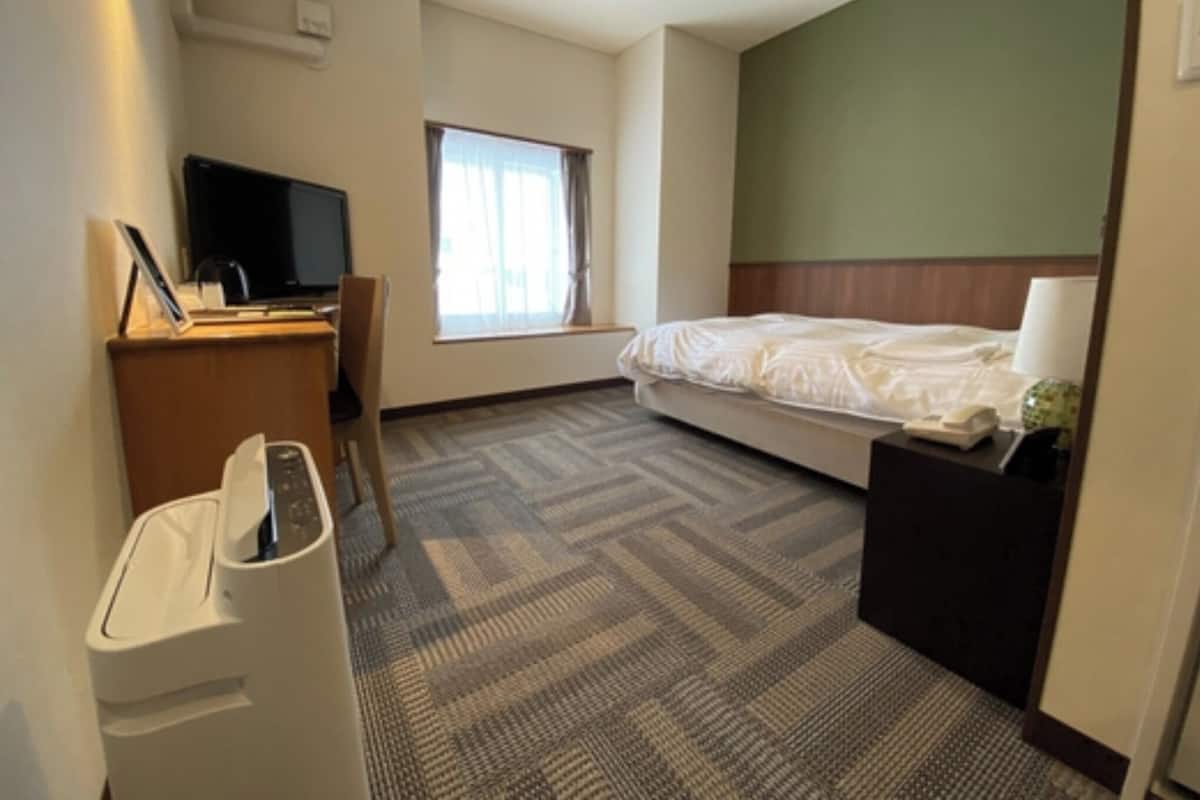 Quarto de hotel com cama, escrivaninha com computador, cadeira, lixeira e janela com cortinas.