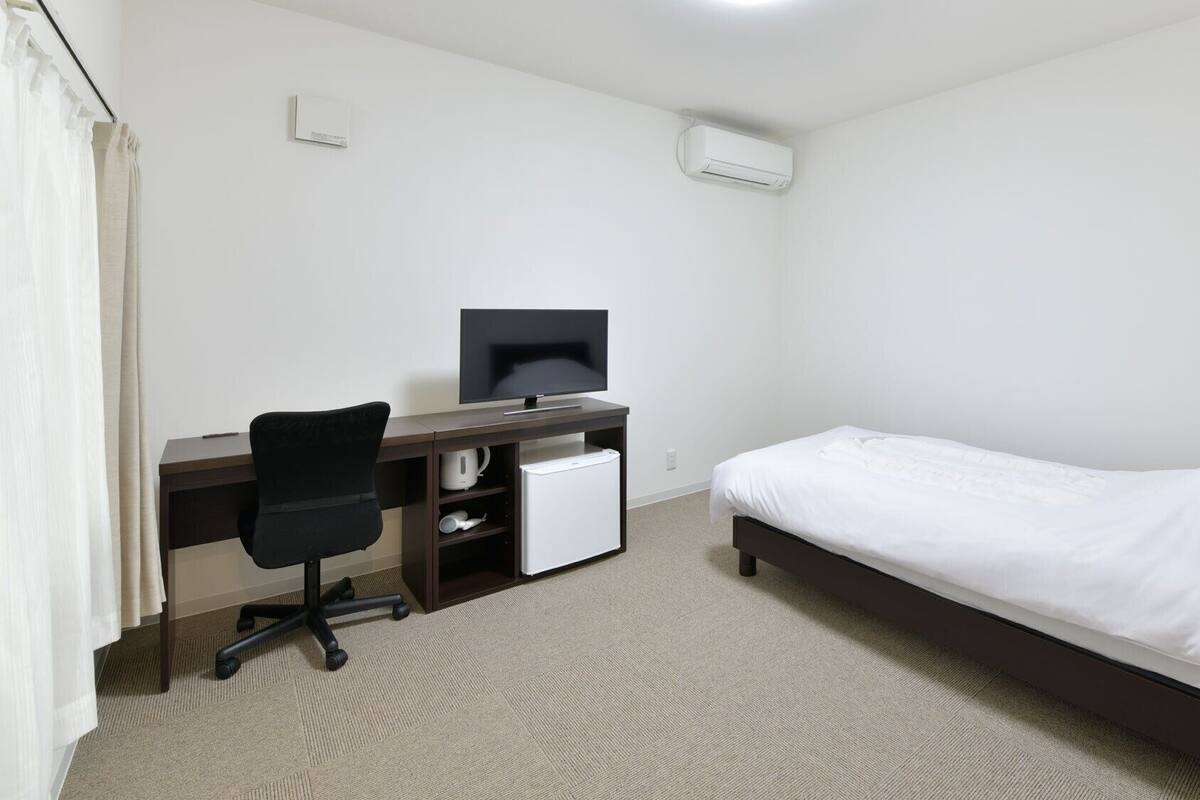 Quarto de hotel com cama, escrivaninha com computador, cadeira, pequeno frigorífico e unidade de ar condicionado.