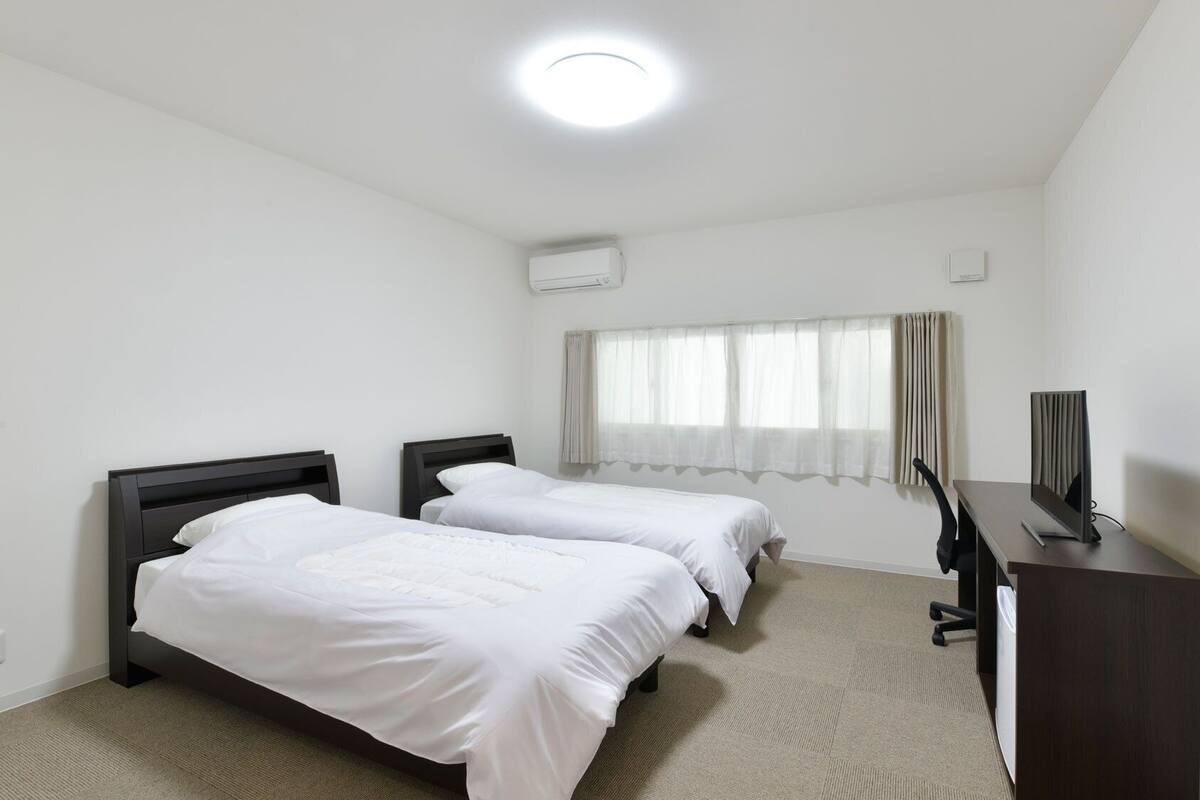 Quarto de hotel com duas camas, uma escrivaninha, uma televisão e uma janela com cortinas.