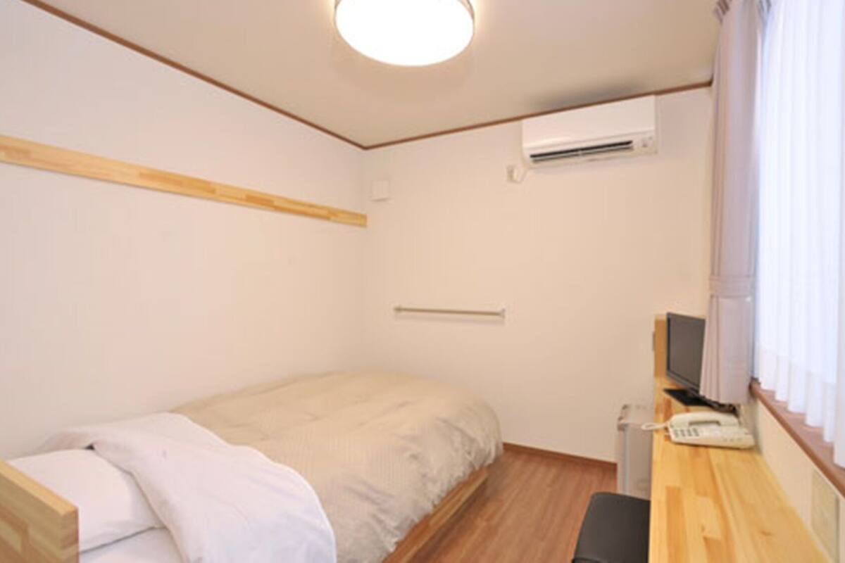 Quarto pequeno e limpo, com uma cama, uma escrivaninha com computador e um ar condicionado.