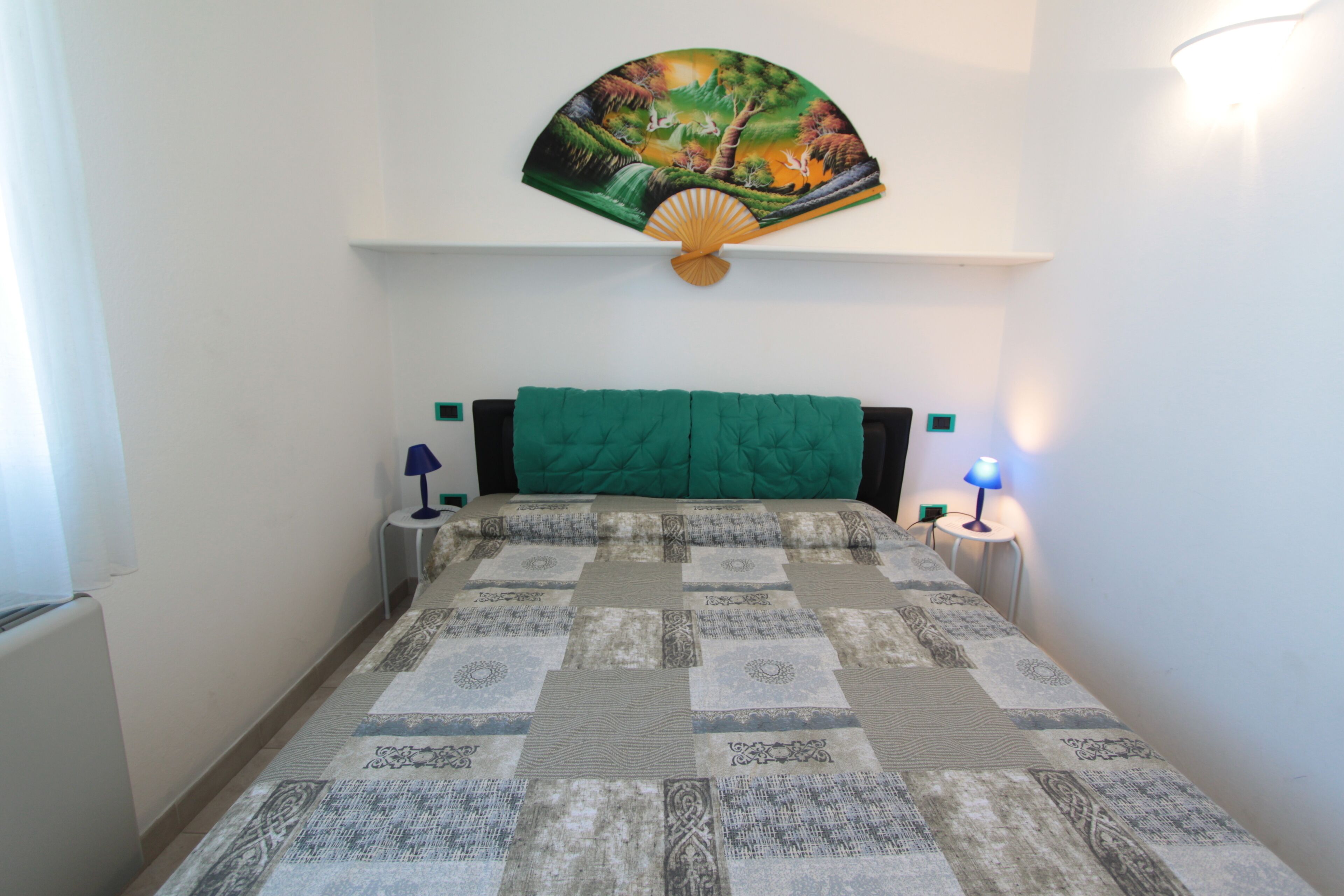 Quarto com cama, cabeceira verde, colcha estampada, ventoinha na parede e abajures laterais.