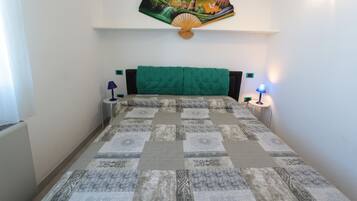 Quarto com cama, cabeceira verde, colcha estampada, ventoinha na parede e abajures laterais.
