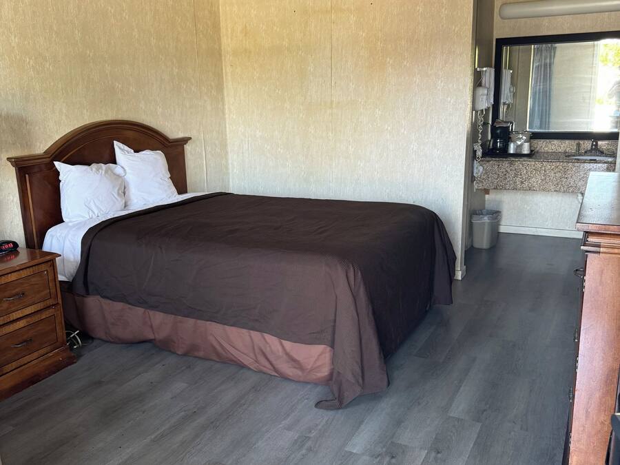 Quarto com cama grande, cabeceira de madeira e cómoda. Há uma pequena escrivaninha e um espelho no canto.