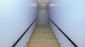 Uma escadaria com degraus bege e corrimãos pretos, que leva a um patamar com um aviso de proibido fumar e um extintor.