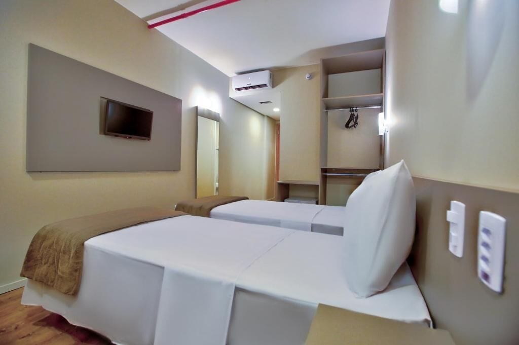 Quarto de hotel com duas camas, uma TV de tela plana na parede, aparelho de ar condicionado e um pequeno nicho com gancho.