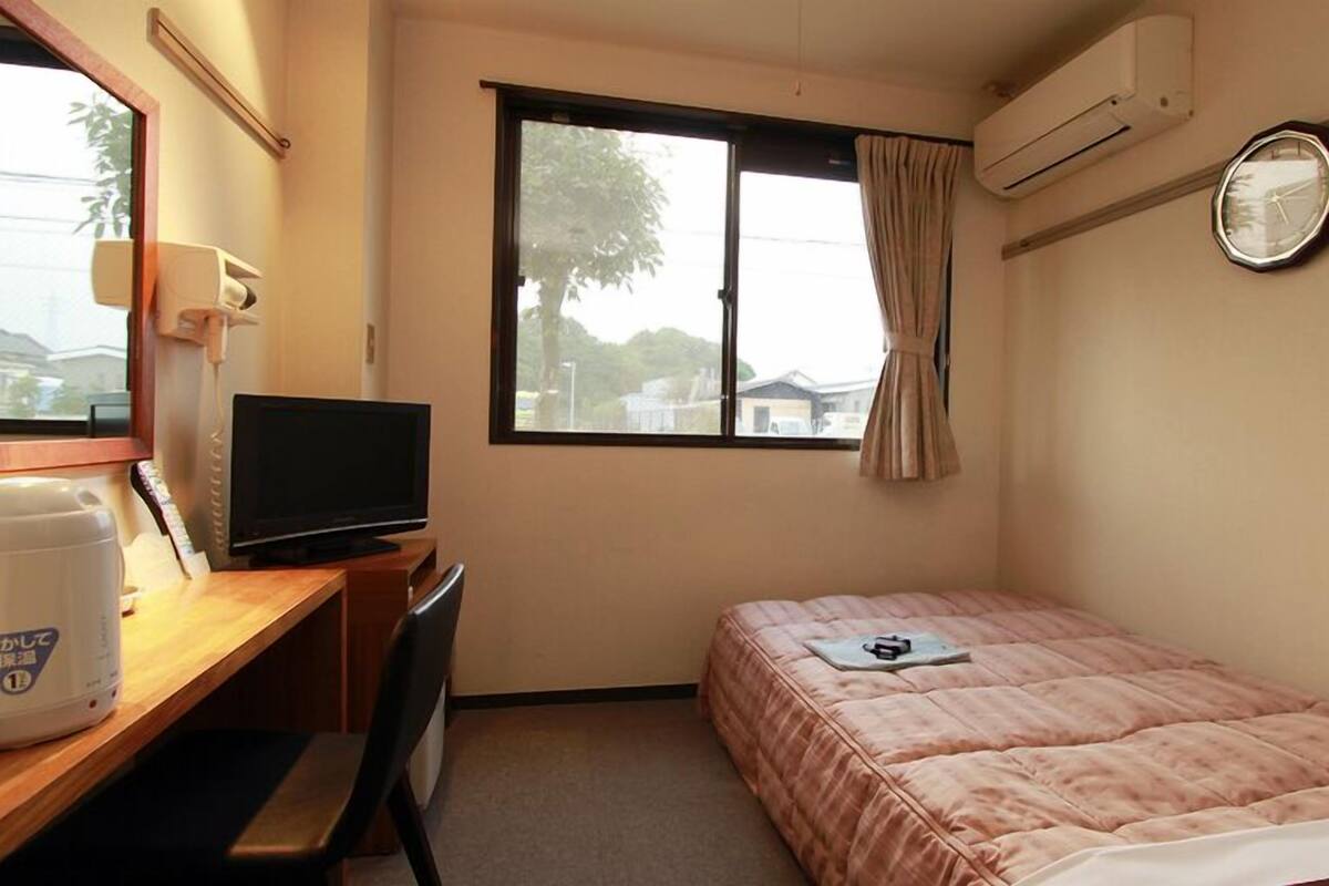 Quarto de hotel com cama, escrivaninha com televisão, janela com cortinas e aparelho de ar condicionado.