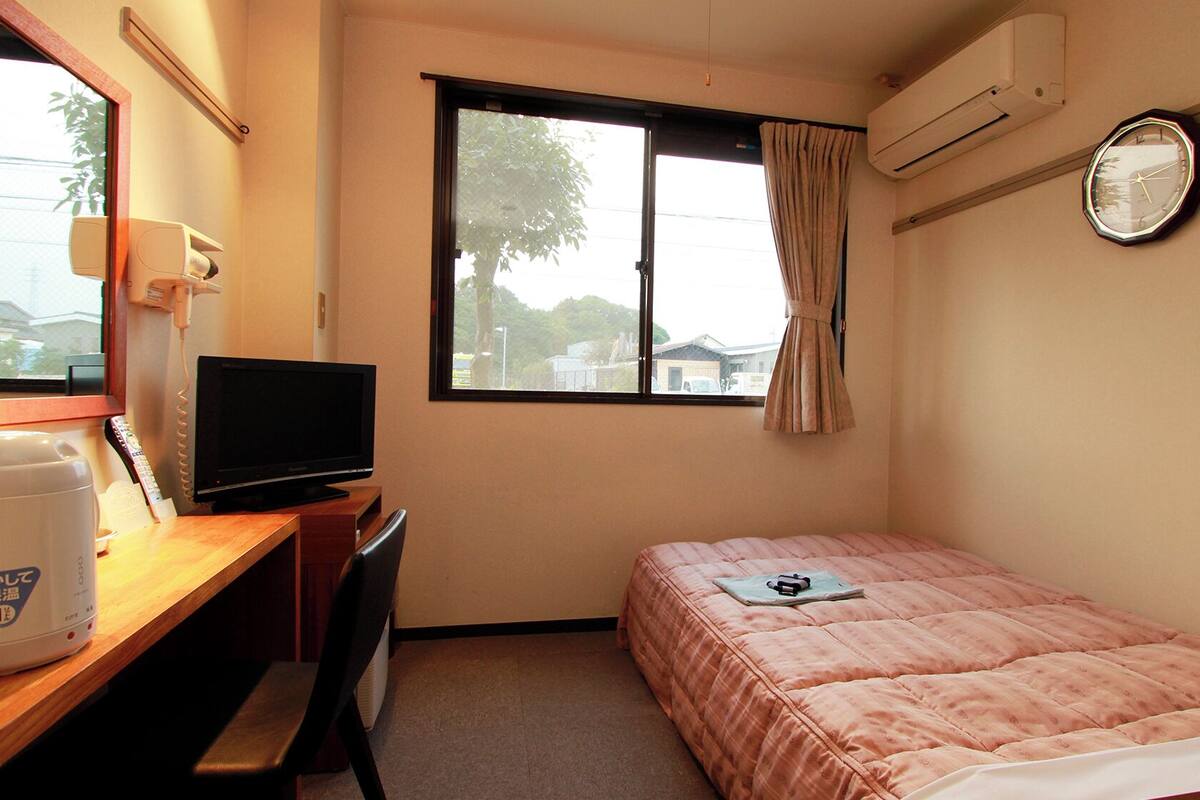 Quarto de hotel com cama, escrivaninha, televisão, máquina de café, janela com cortinas e ar condicionado.