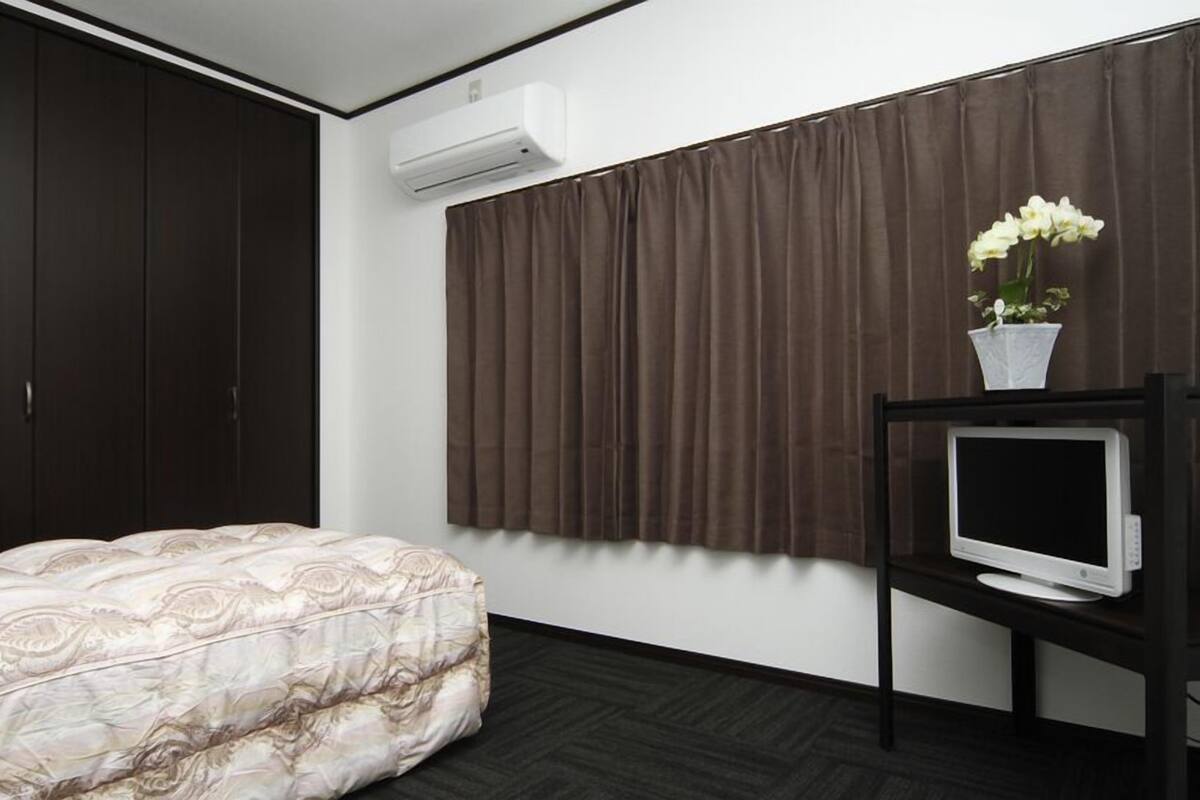 Quarto com cama, guarda-roupa escuro, ar condicionado, cortinas, uma mesinha com TV e um vaso de planta.