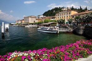 Uma cena à beira-mar com um barco atracado num cais, rodeado por edifícios e um passeio decorado com flores.