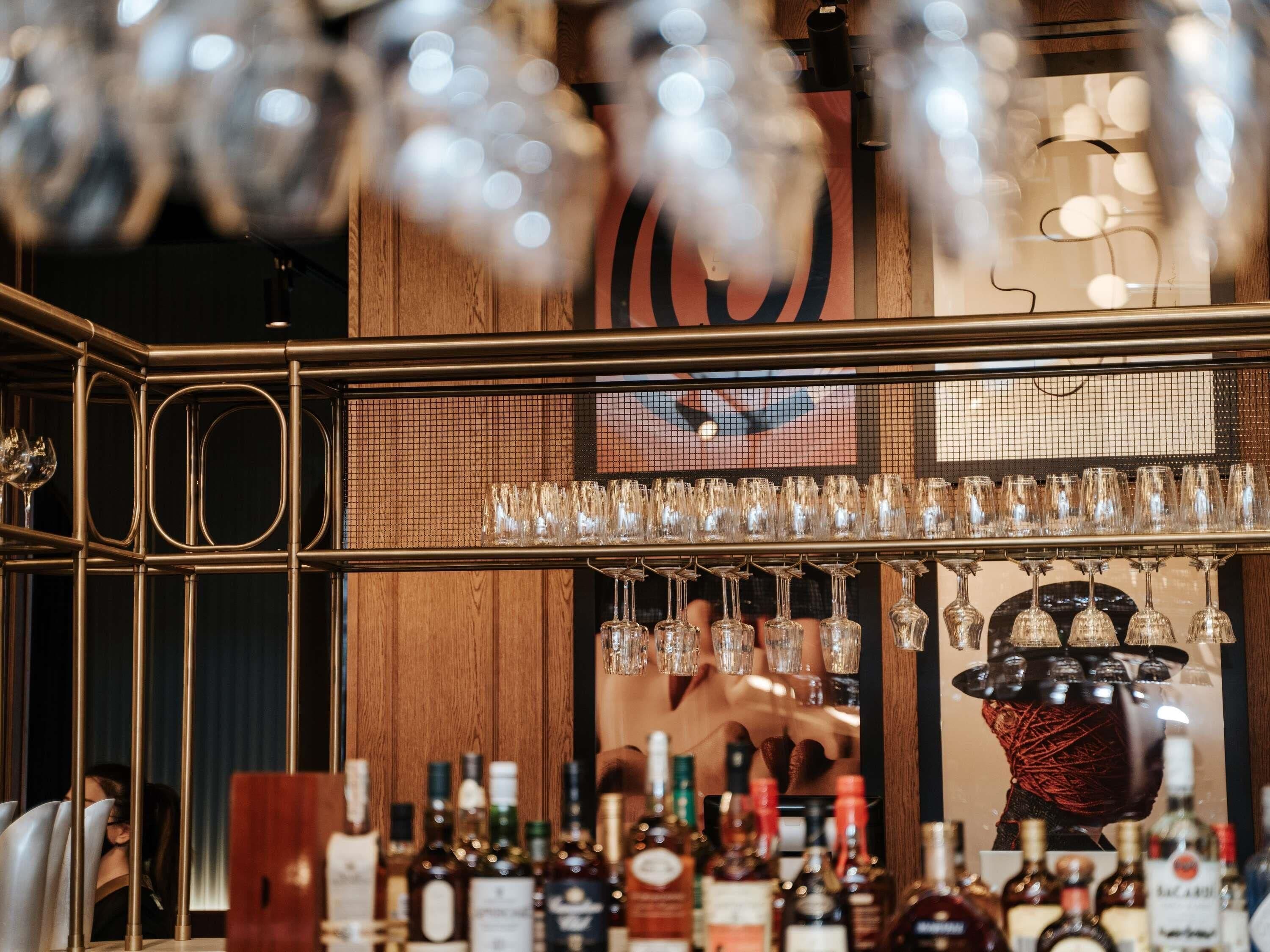 Uma área de bar com painel de madeira, balcão de metal e diversos frascos de bebidas alcoólicas.