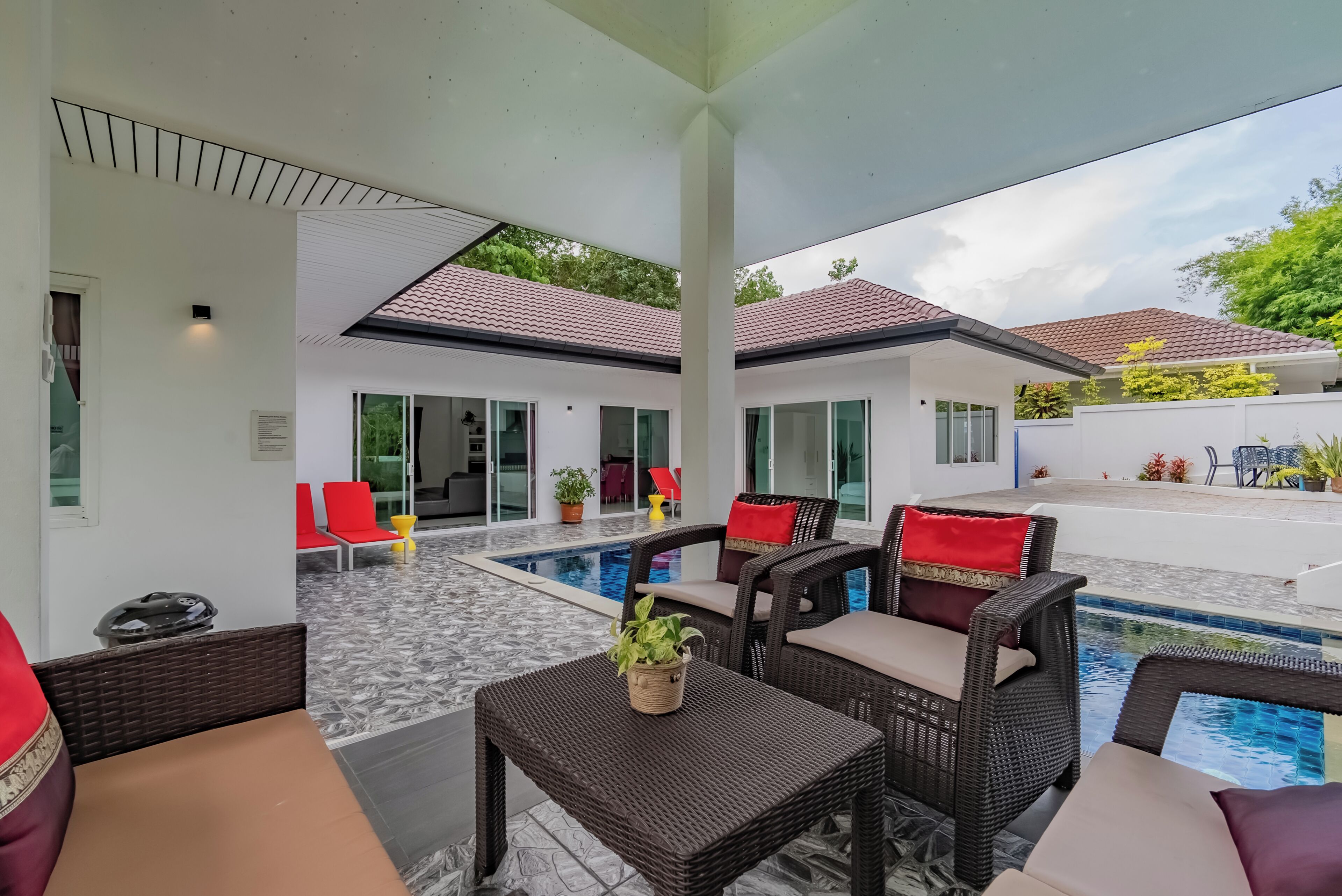 Área de piscina ao ar livre com mobiliário de vime e almofadas vermelhas, um alpendre coberto e uma casa ao fundo.