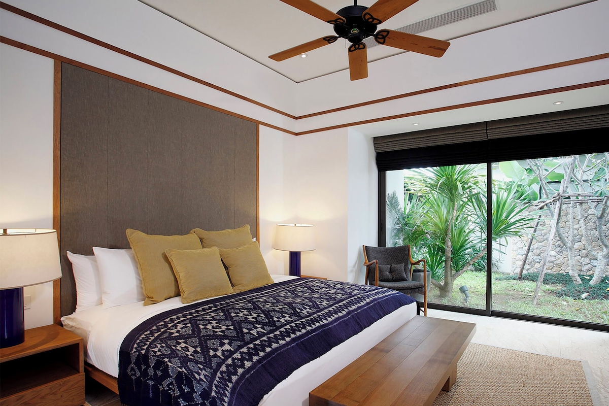 Quarto com cama, mesas de cabeceira, ventilador de teto, um banco e vista para um jardim.