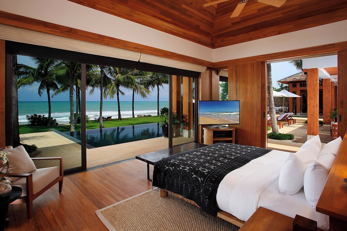 Quarto com uma janela ampla para uma praia, uma cama, uma televisão e teto de madeira.