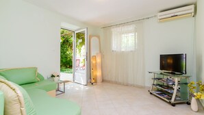 Sala de estar com um sofá verde, uma TV em um suporte e uma porta de vidro deslizante que dá para um jardim.
