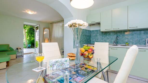Uma cozinha moderna com armários verde claro, uma área de refeições com mesa de vidro, cadeiras brancas e um vaso com flores.