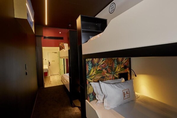 Quarto com beliche, cabeceira estampada com folhagem tropical, travesseiro branco com logotipo e porta que dá para outro quarto.