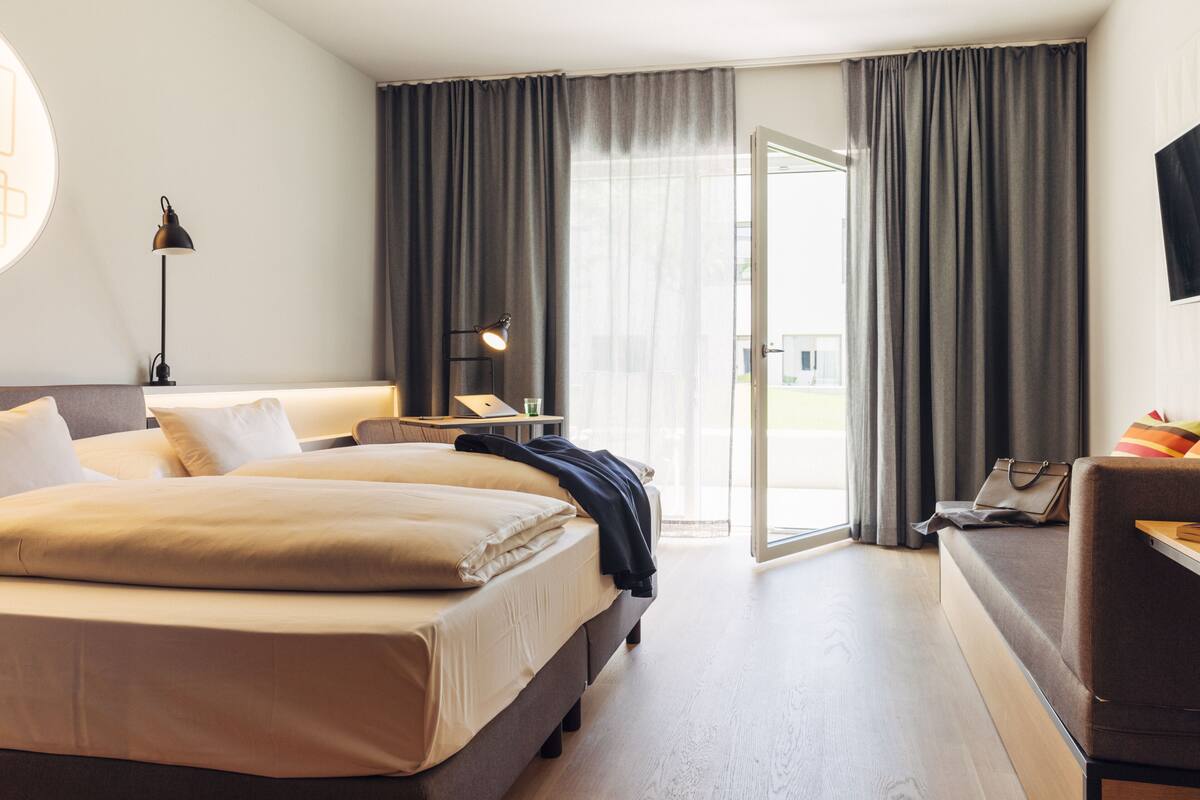 Quarto de hotel com uma cama grande, uma escrivaninha, uma cadeira e um sofá. Há uma janela com cortinas e uma porta que dá para o exterior.