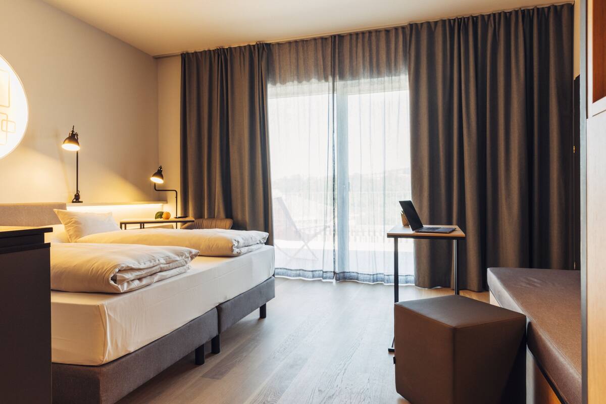 Quarto de hotel com duas camas, uma mesa, um laptop e vista da cidade por grandes janelas.