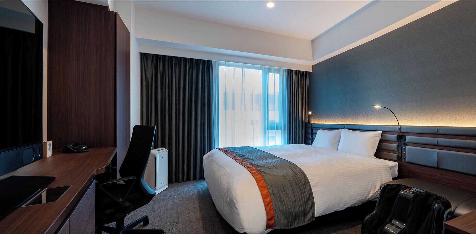 Quarto de hotel com cama, escrivaninha, cadeira e uma janela grande com cortinas.