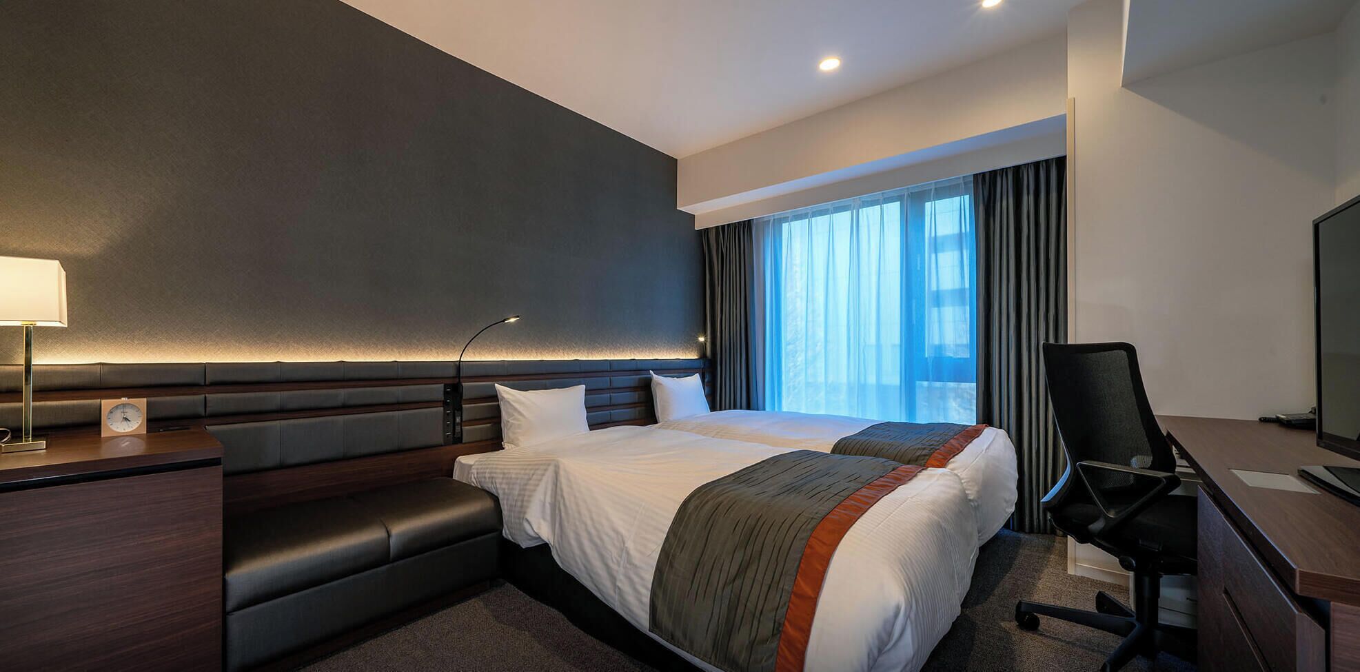 Quarto de hotel moderno com uma cama grande, escrivaninha, cadeira, televisão e janela com cortinas.
